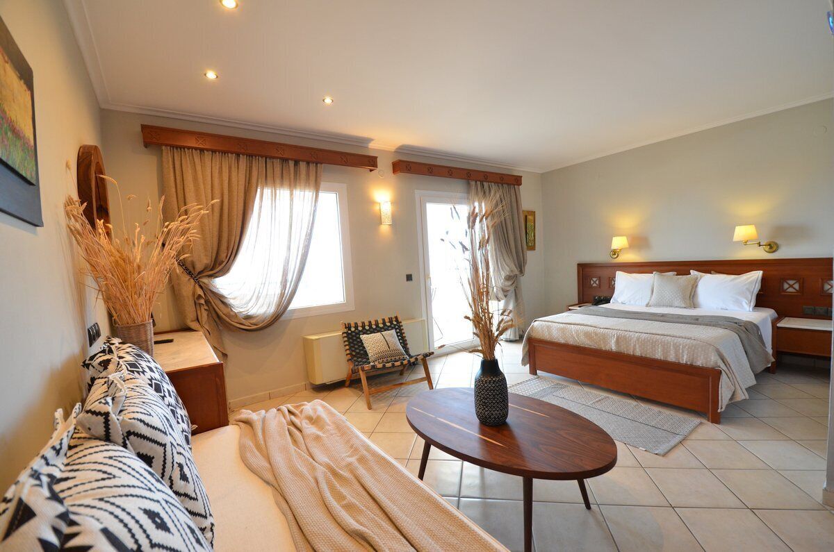 Quarto de hotel com cama, sofá, mesa e cadeira. Cortinas, um vaso com plantas secas e arandelas na parede.