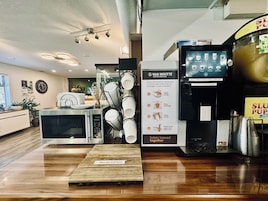 Interior de uma cafeteria com uma máquina de café, um dispensador de bebidas quentes e um aviso detalhando os passos do serviço de café.