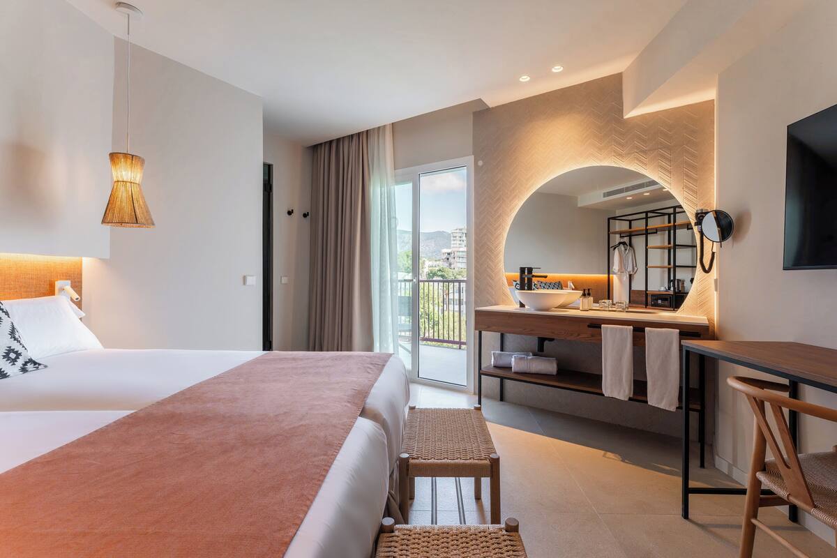 Quarto de hotel moderno com uma cama grande, uma escrivaninha com espelho, um banheiro com pia e uma varanda com vista.