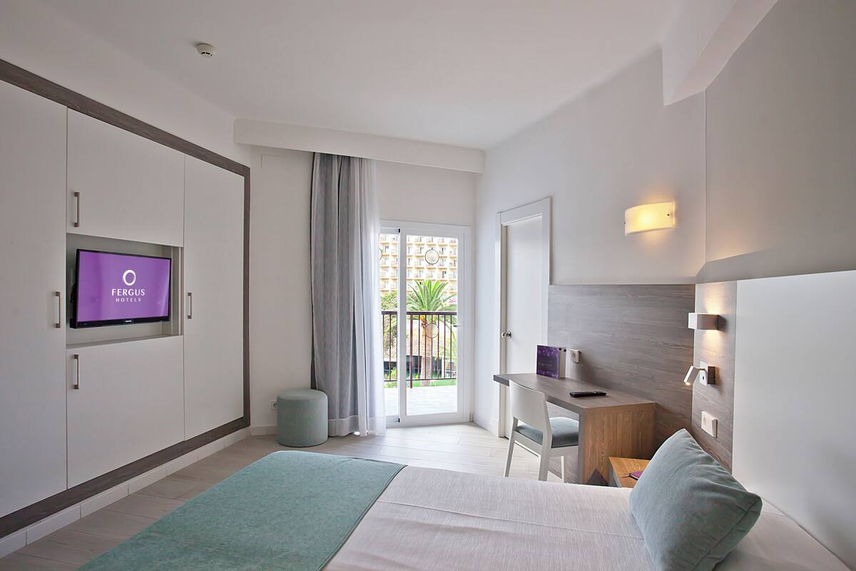 Quarto de hotel moderno com cama, escrivaninha, televisão e varanda com vista para o verde.