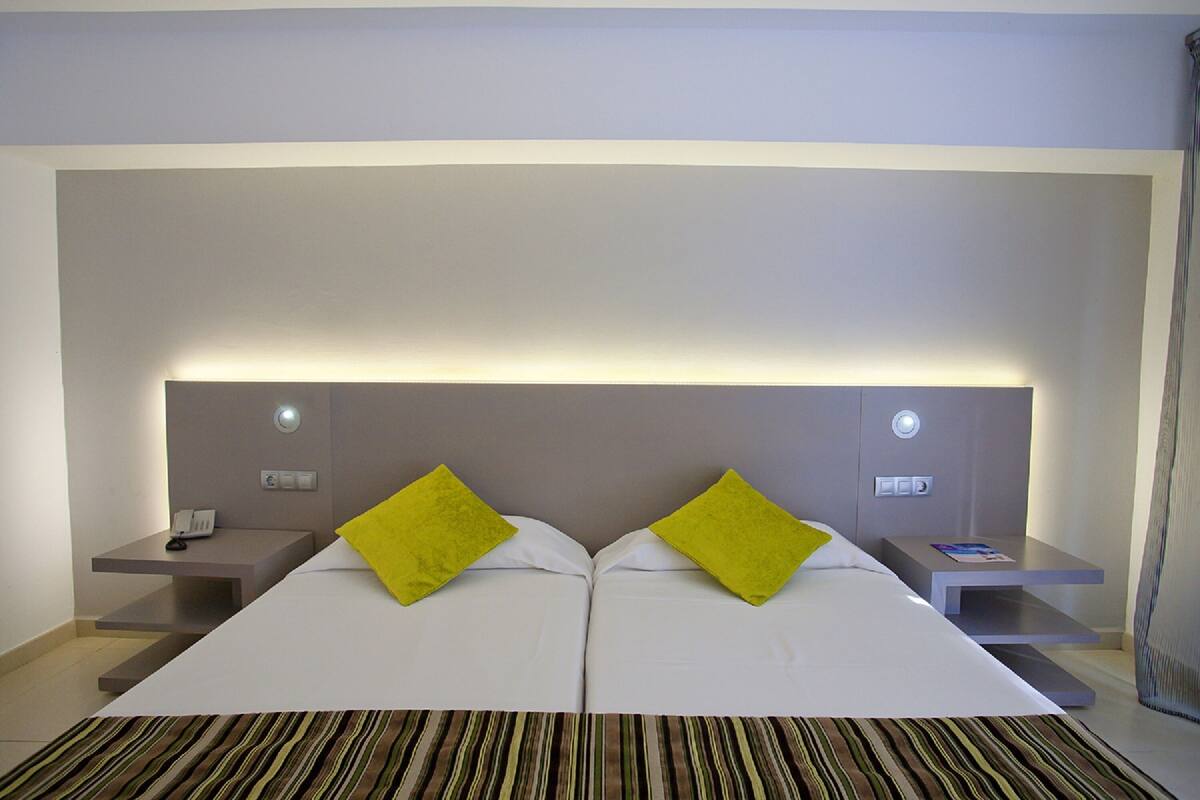 Uma cama de casal com duas almofadas, uma mesa de cabeceira com um telefone e uma mesinha de apoio com um livro.