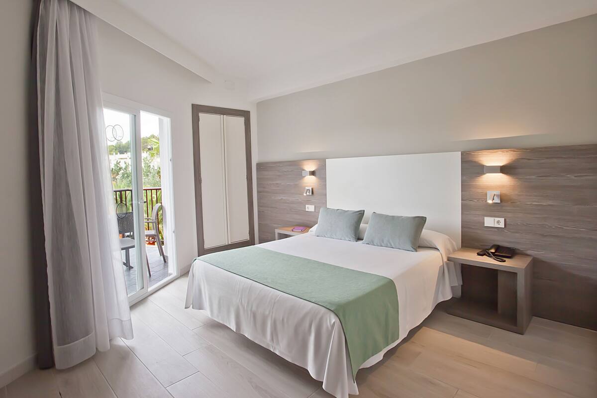 Quarto moderno com cama grande, mesa de cabeceira, luminária fixada na parede e porta de vidro deslizante para uma varanda.