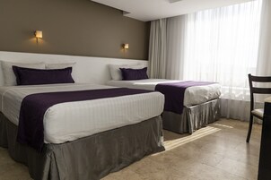 Quarto de hotel com duas camas, uma cadeira e uma janela grande com cortinas.