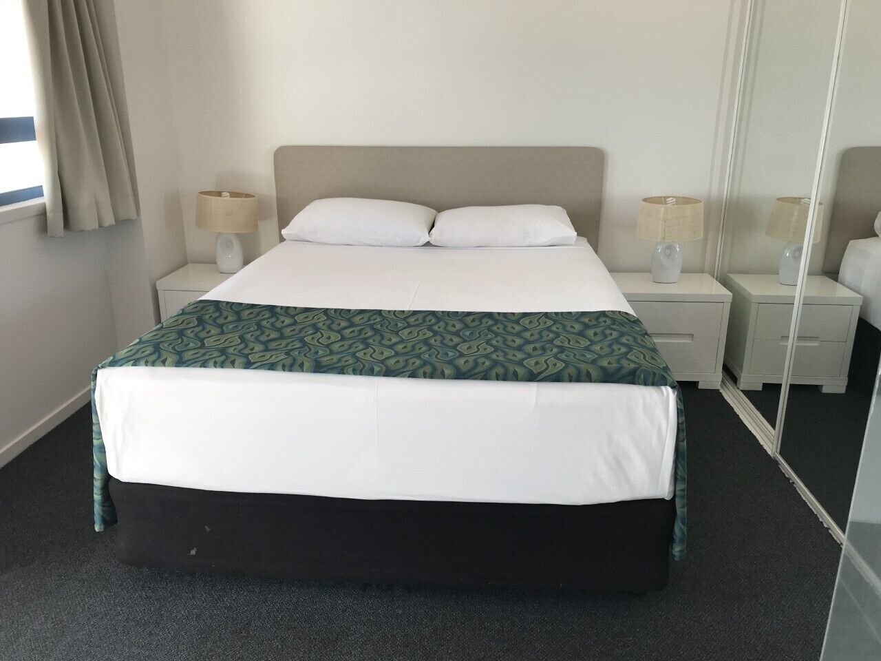 Quarto de hotel com cama, duas mesas de cabeceira com luminárias, espelho e janela com cortinas.