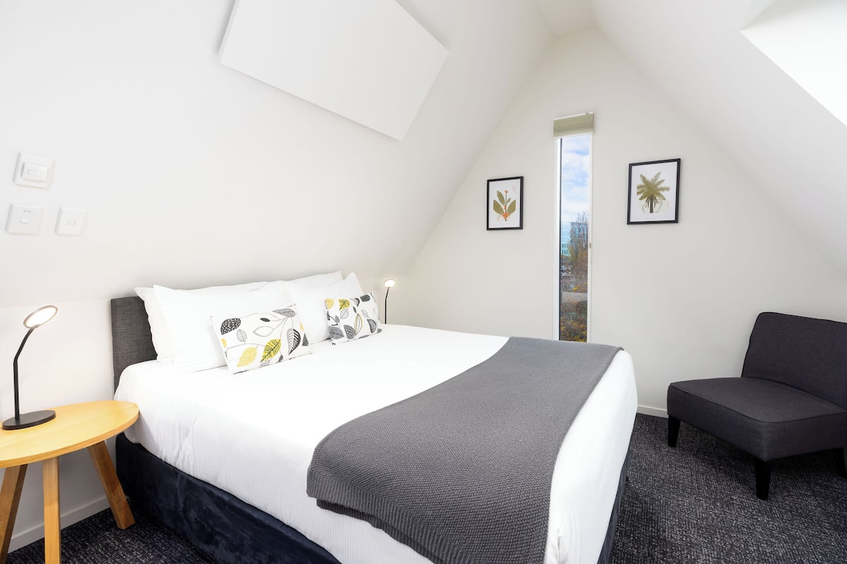 Quarto de hotel moderno com uma cama grande, uma mesa de trabalho, uma cadeira e vista para o exterior.