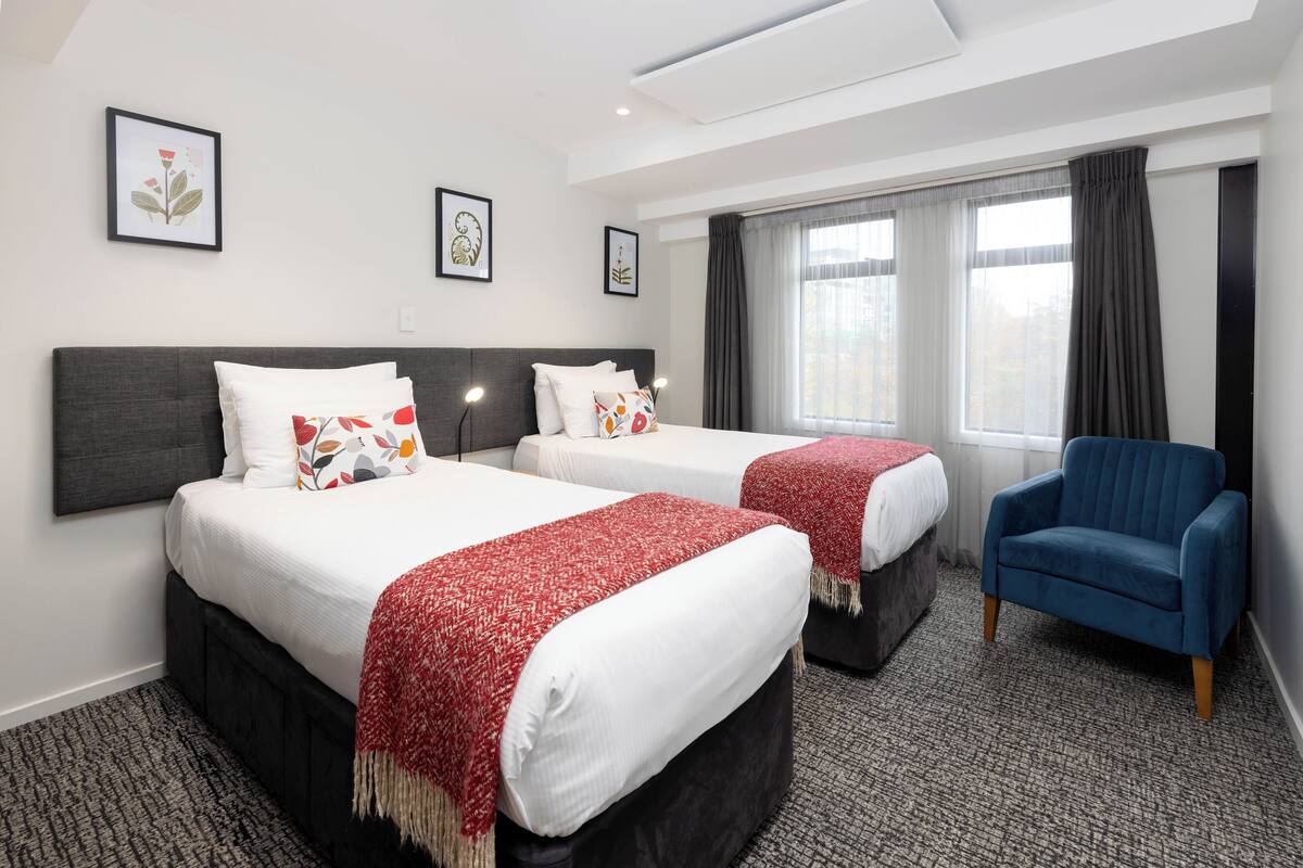 Quarto de hotel com duas camas, uma poltrona azul e janelas amplas com cortinas.