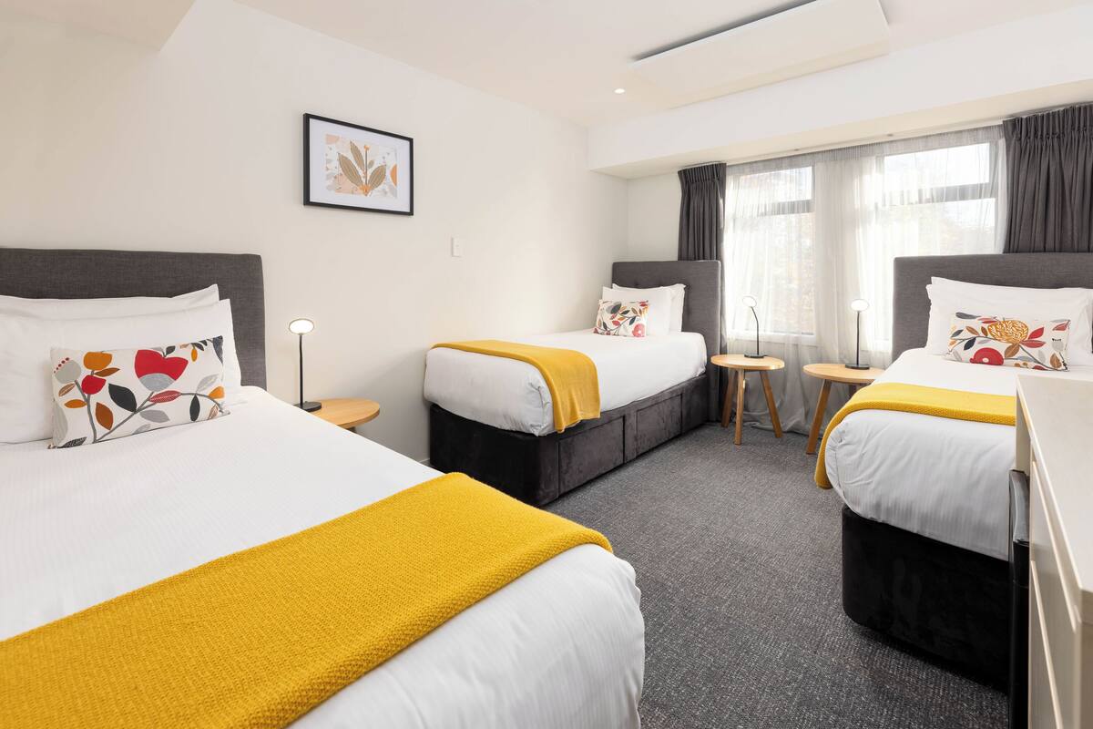 Quarto de hotel com duas camas, cada uma com um edredom branco e um cobertor amarelo. Há duas mesas de cabeceira com luminárias, um pequeno banquinho e um quadro na parede.