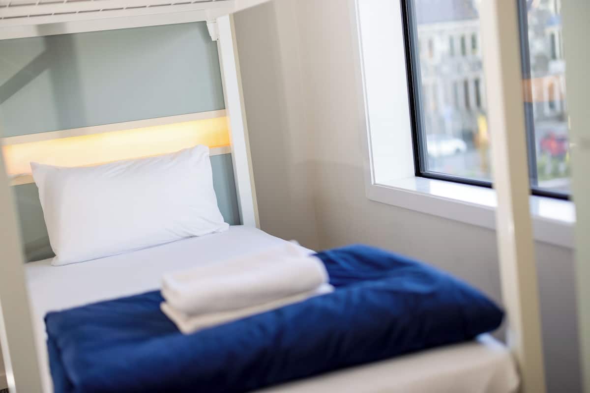 Uma cama de solteiro com travesseiro branco e roupa de cama azul, uma janela com vista para edifícios e cabeceira branca.
