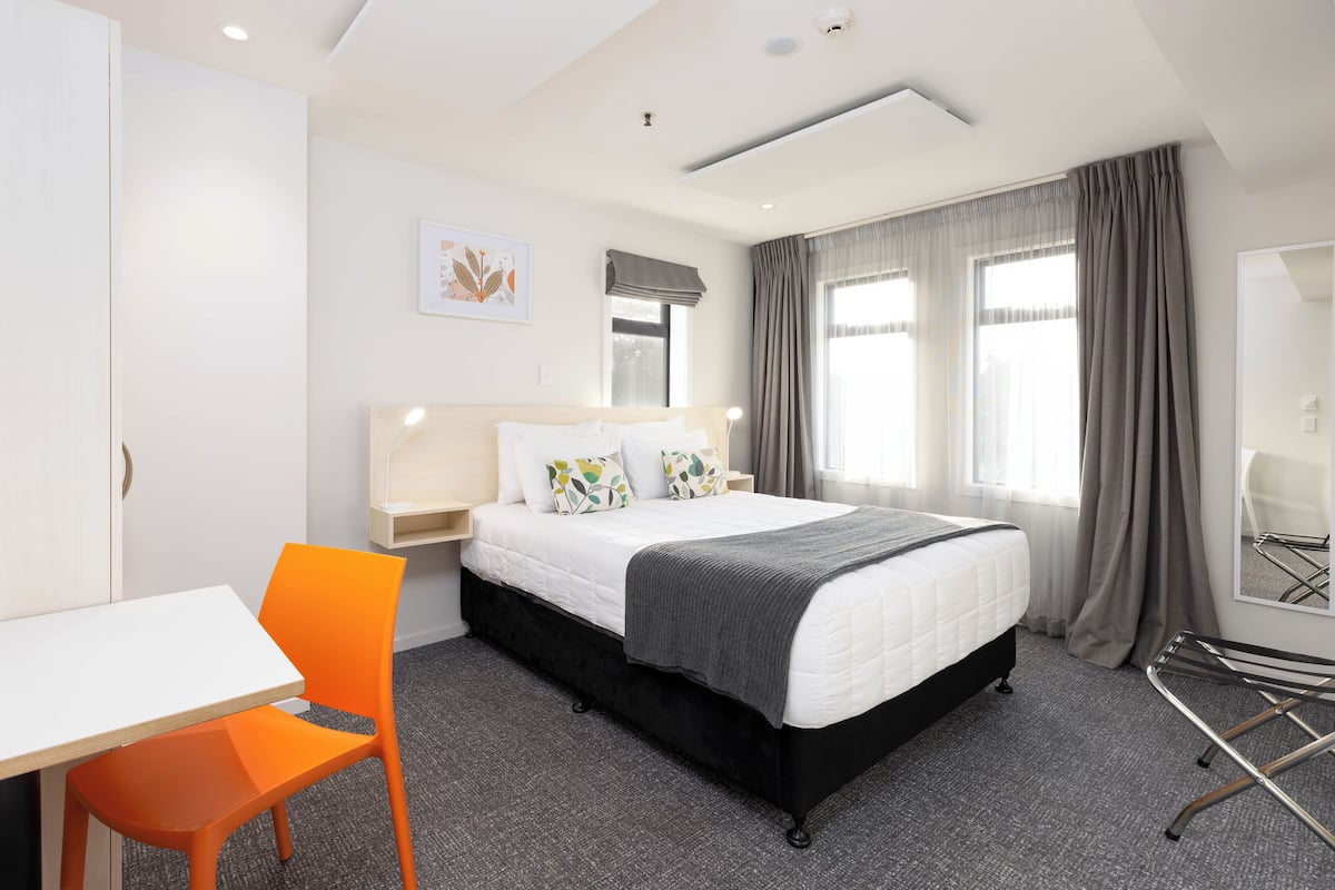 Quarto de hotel com uma cama, uma escrivaninha, uma cadeira laranja, um tapete cinza e uma janela com cortinas.