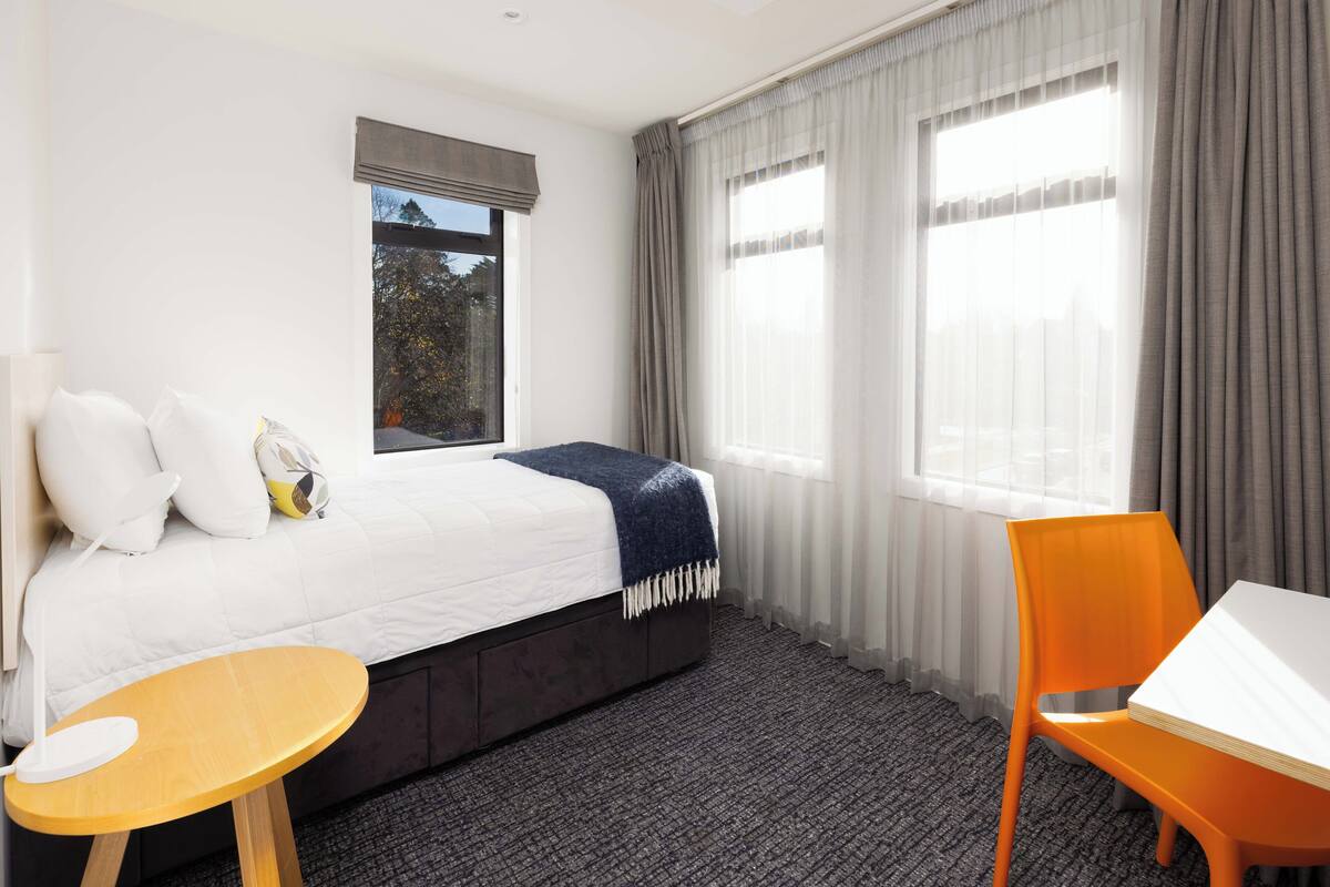 Quarto de hotel com cama, uma mesinha, cadeira laranja e janelas amplas com cortinas.