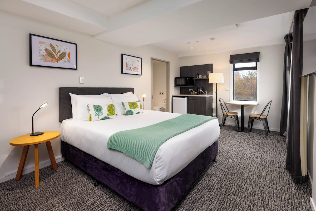 Quarto de hotel com cama, secretária, cadeira, pequena cozinha e janela.