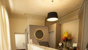 Uma área de recepção de hotel com um balcão de mármore, um espelho redondo, um relógio e um vaso com flores.