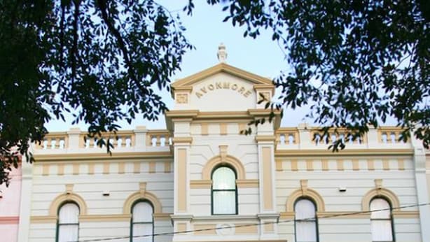 Um edifício histórico com entrada central, varandas de dois andares e uma placa com a inscrição
