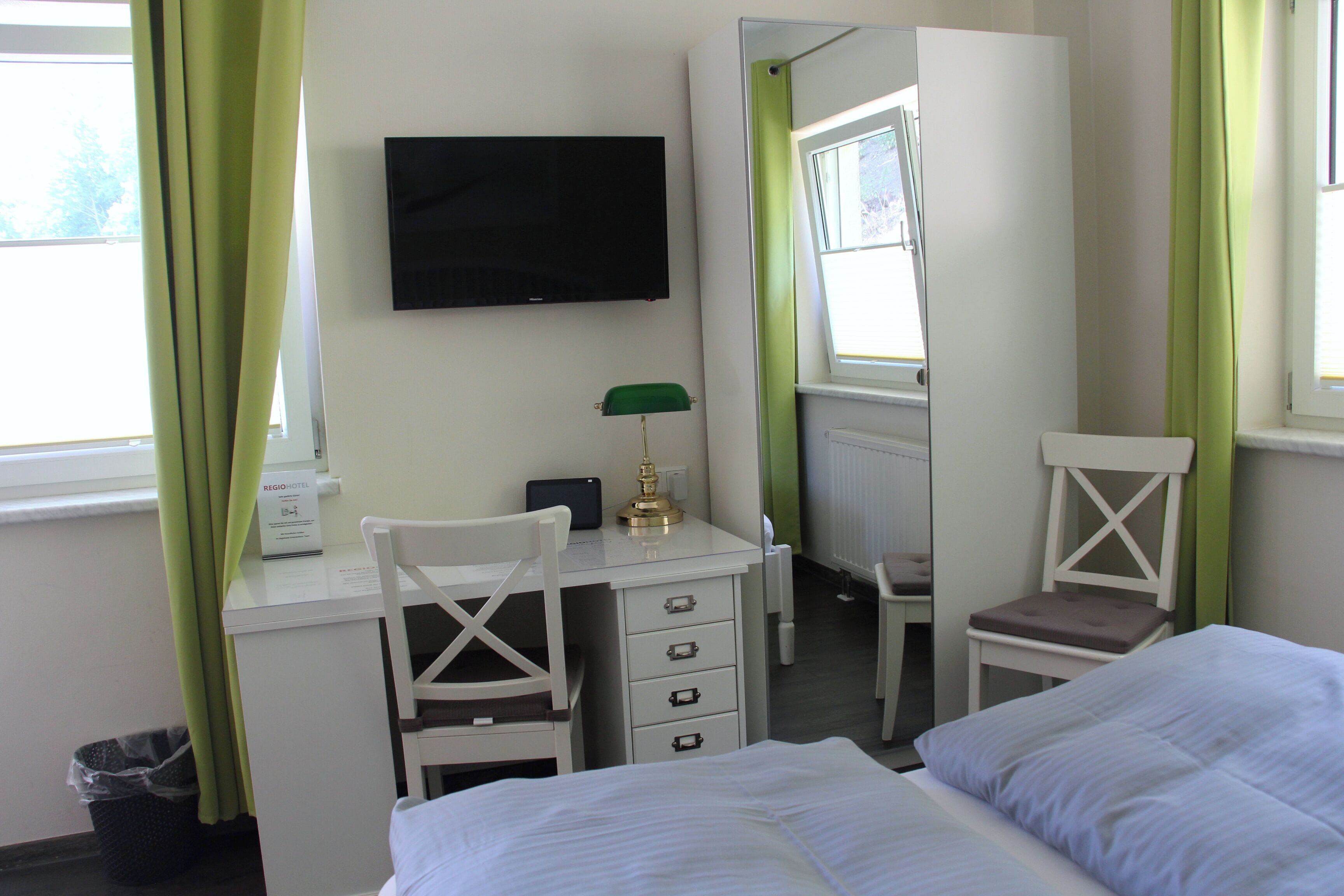 Quarto de hotel com escrivaninha, cadeira, cama, televisão e janela com cortinas.
