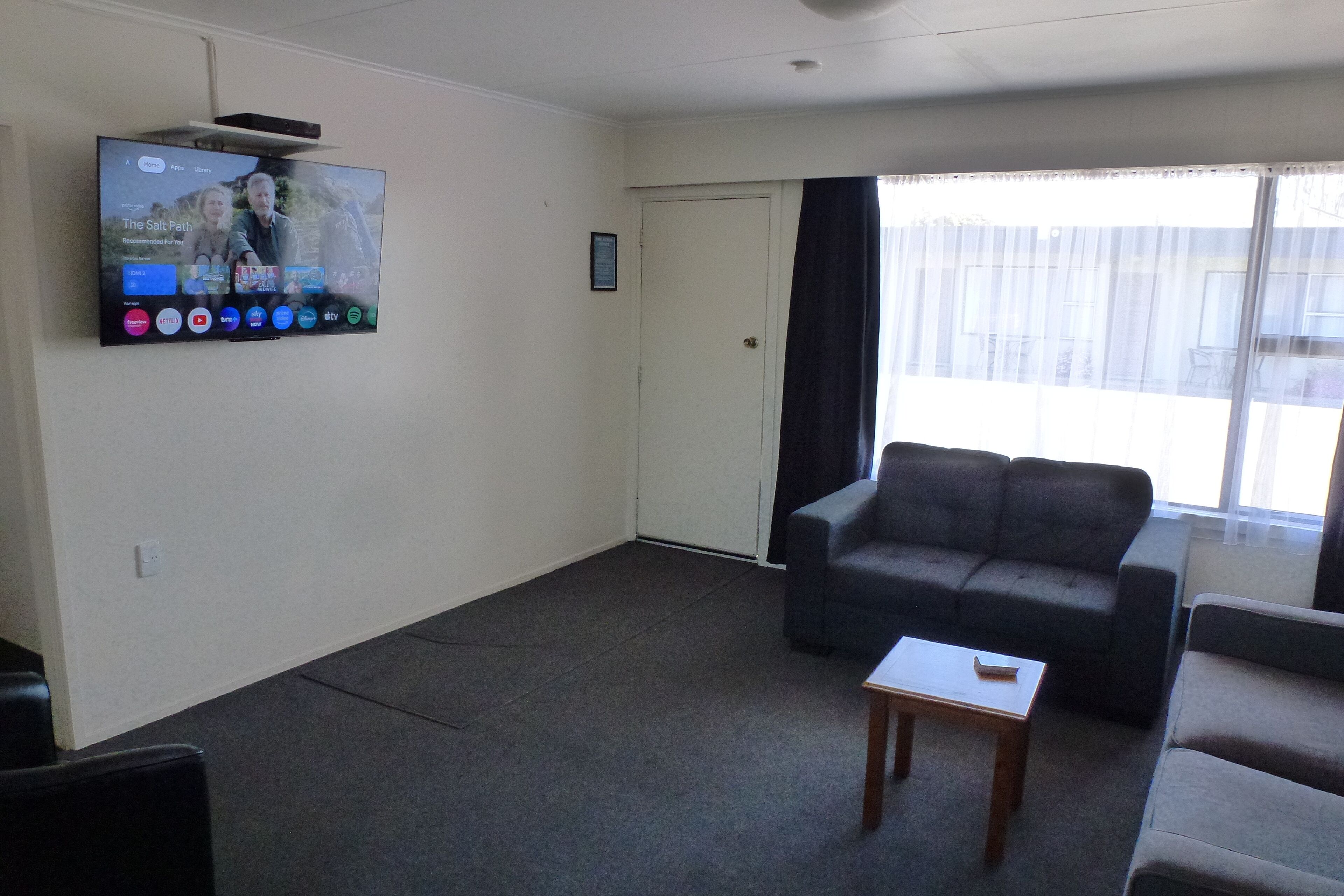 Sala de estar com televisão de tela plana na parede, sofá cinza, uma pequena mesa de madeira e uma janela com cortinas.