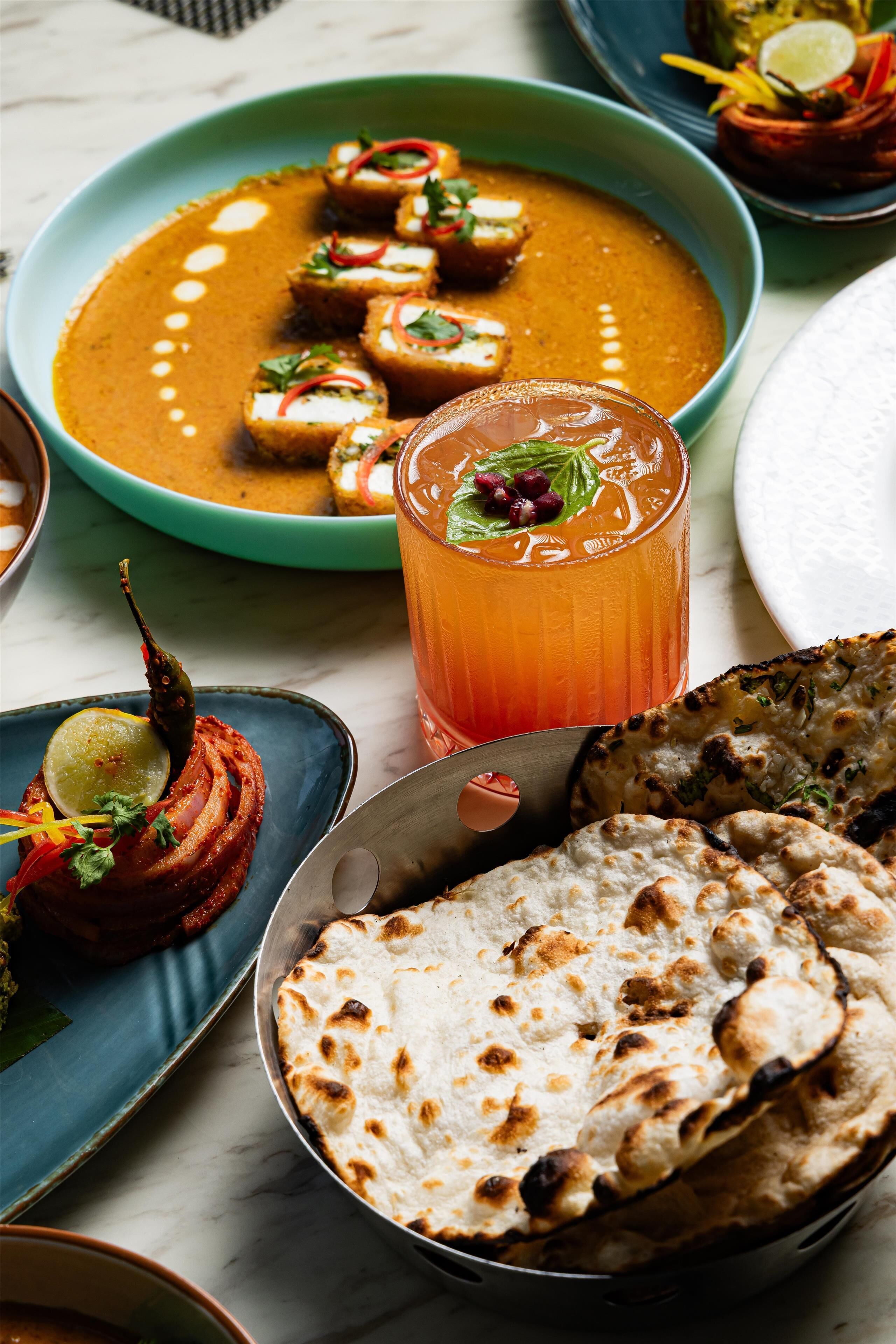 Uma mesa com diversos pratos, incluindo um tigela de curry, pão naan e um copo de bebida de laranja.