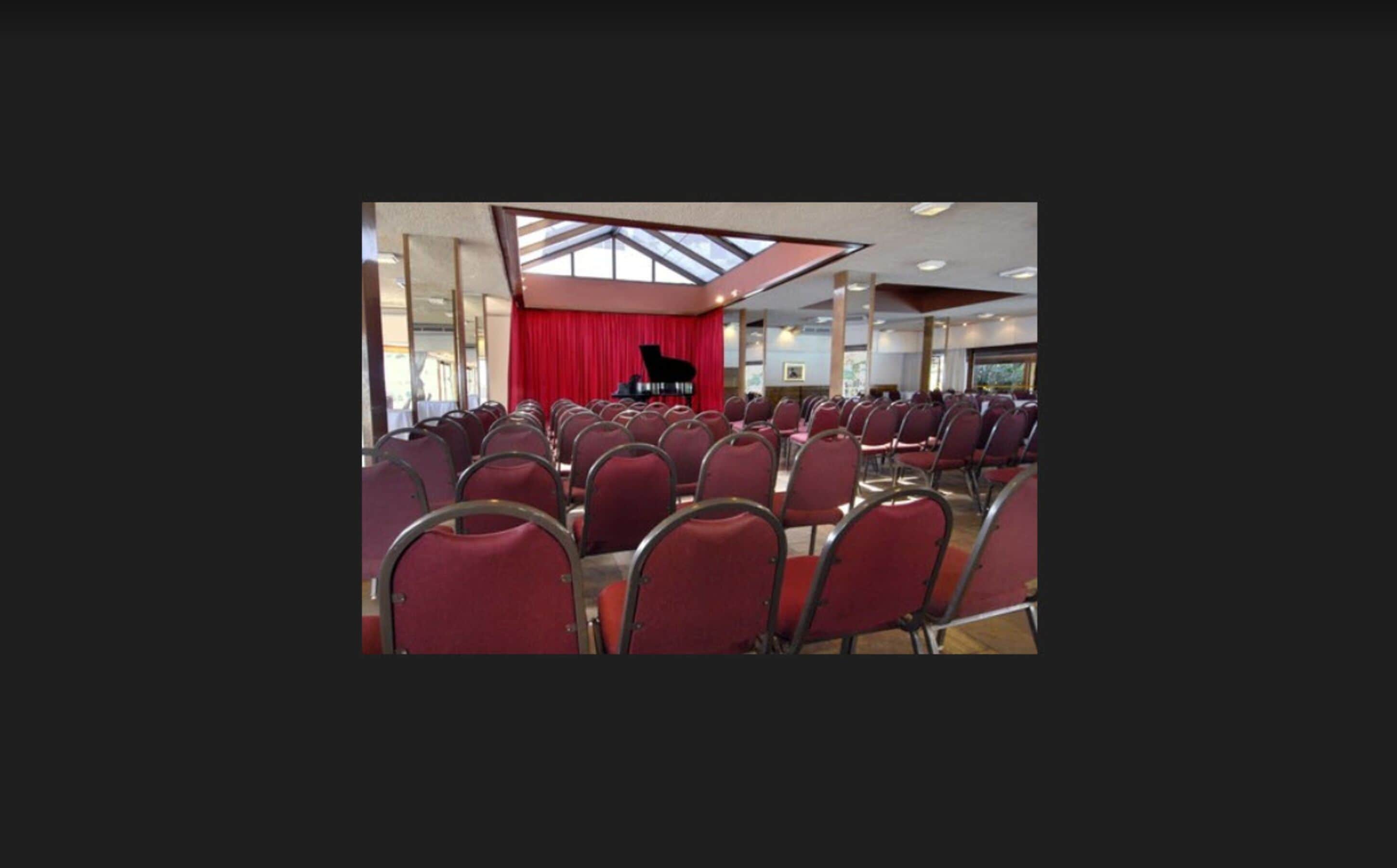 Uma sala de conferências com fileiras de cadeiras vermelhas, um palco com um piano e janelas amplas.