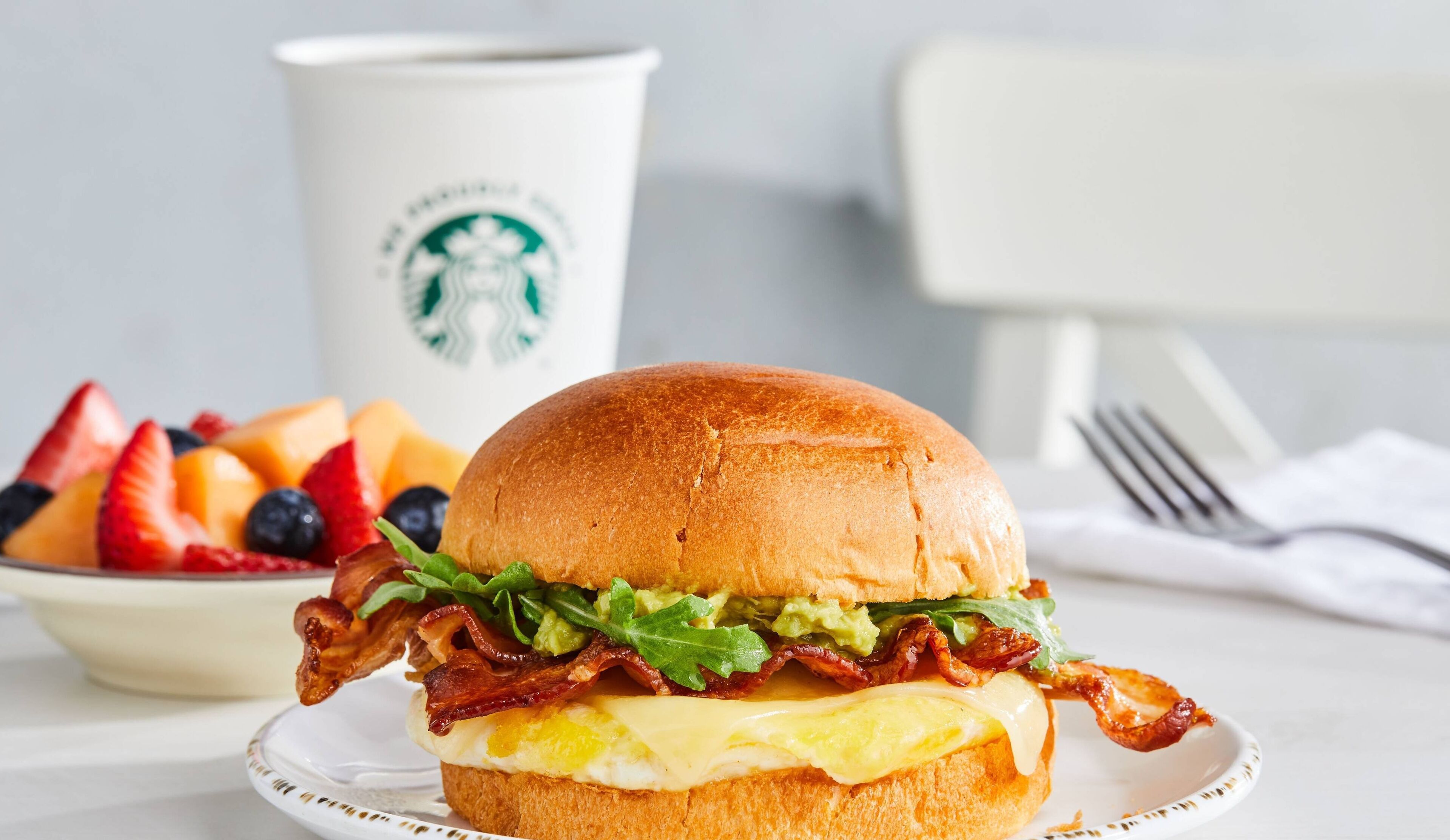 Um sanduíche de café da manhã com bacon, abacate e ovo no pão, um prato de frutas e uma xícara da Starbucks ao fundo.