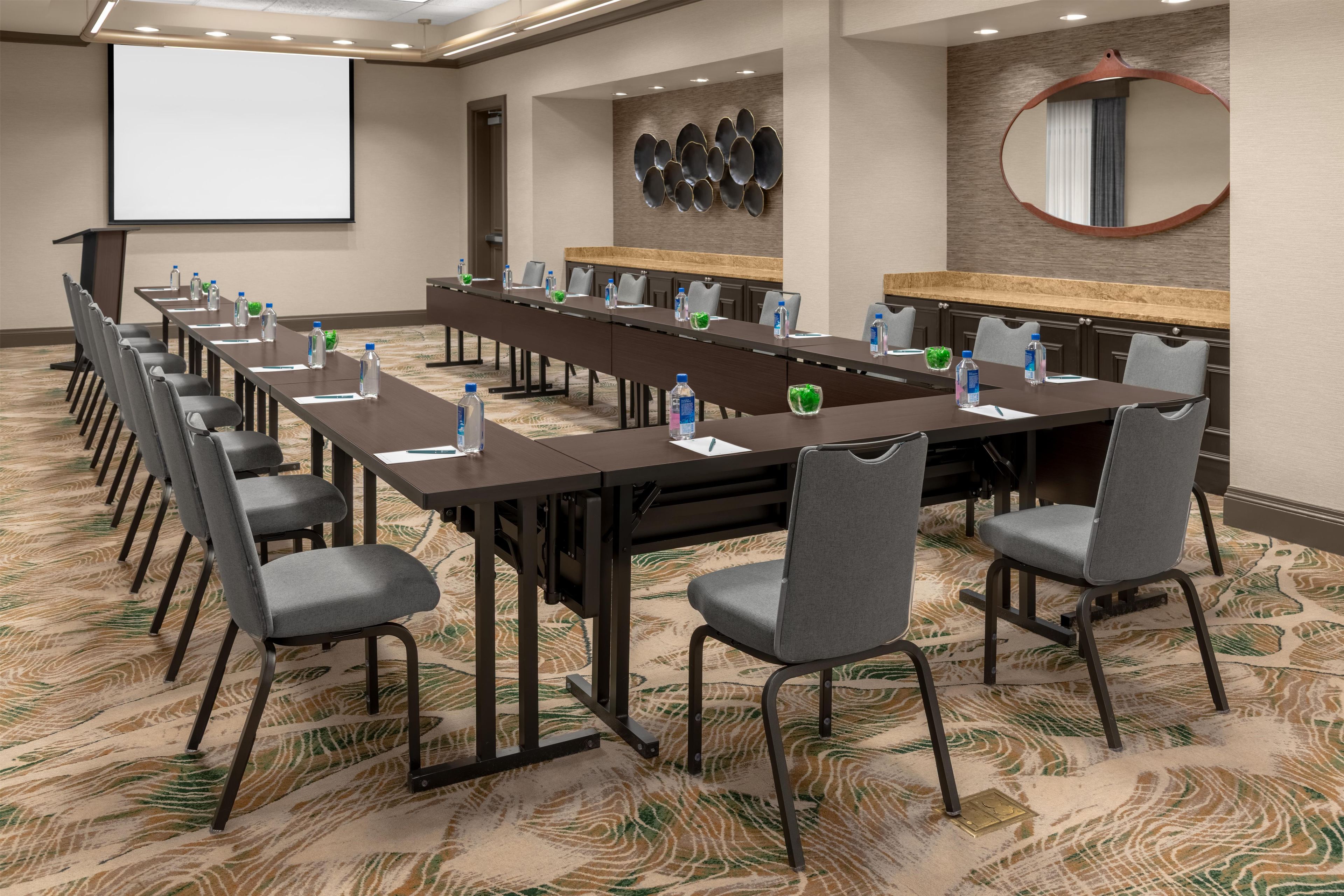 Uma sala de conferência com uma mesa comprida, cadeiras, garrafas de água e uma tela de projetor.