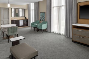 Quarto de hotel com cozinha compacta, área de refeições com mesa e cadeiras, espaço de estar com duas poltronas, uma janela grande com cortinas e uma televisão.