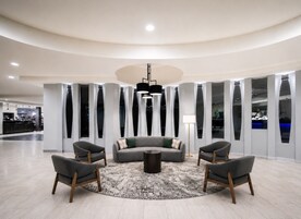 Um lobby moderno com uma mesa de centro redonda, sofás cinzas e cadeiras de madeira.