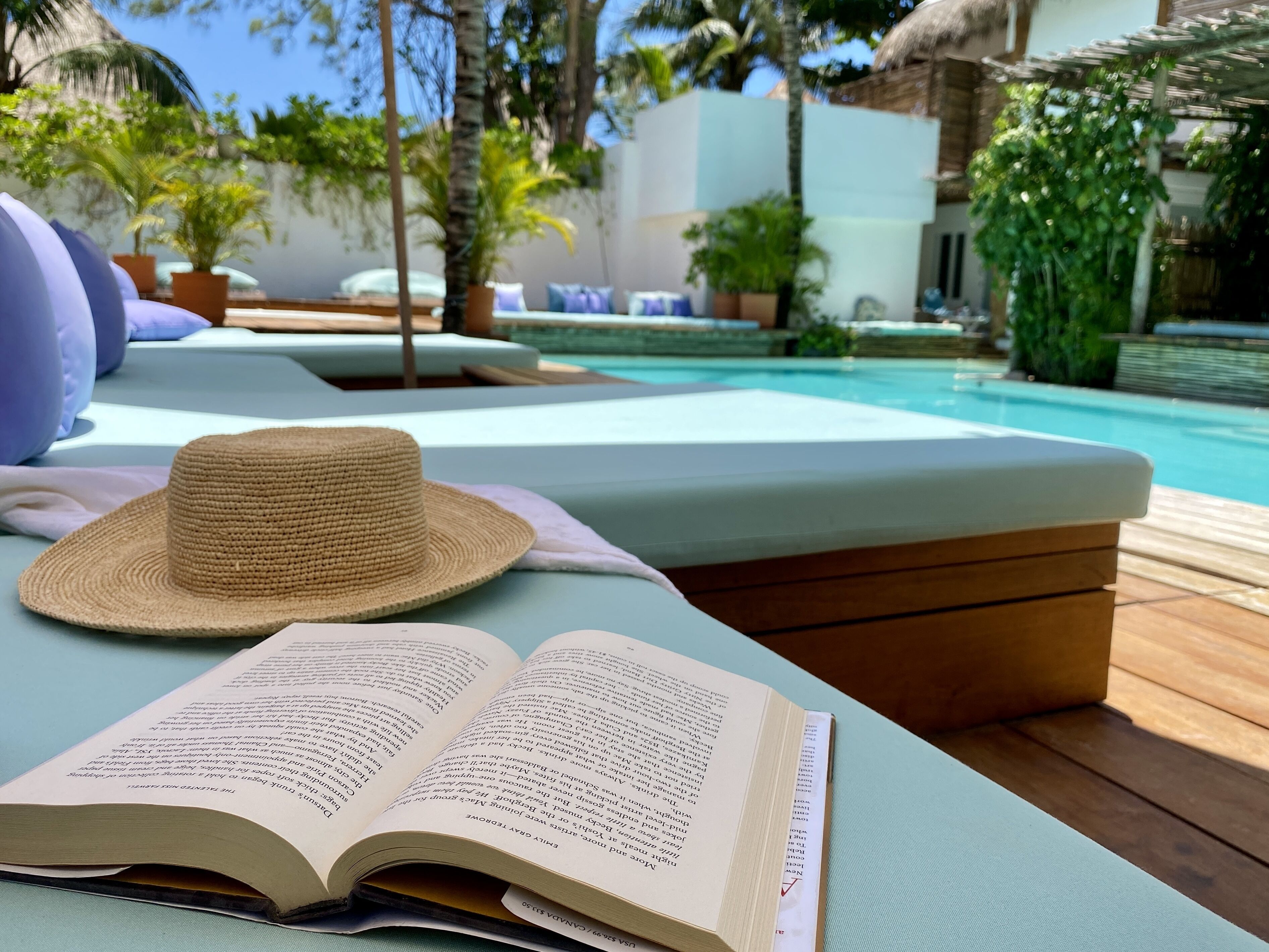 Uma área de lounge à beira da piscina, com um chapéu de palha e um livro aberto sobre um banco de madeira.