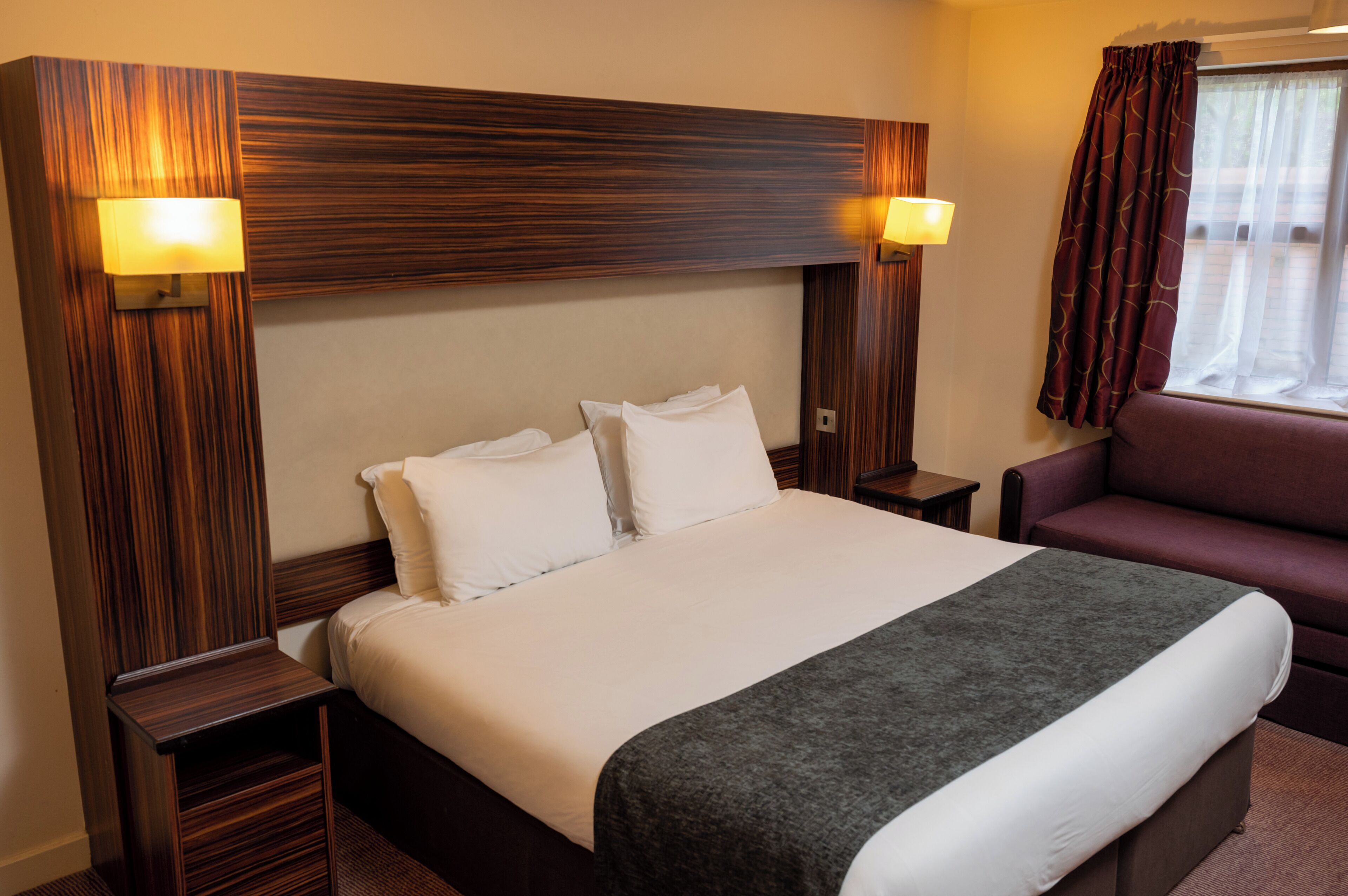 Quarto de hotel com cama grande, duas mesas de cabeceira, um sofá e janela com cortinas.