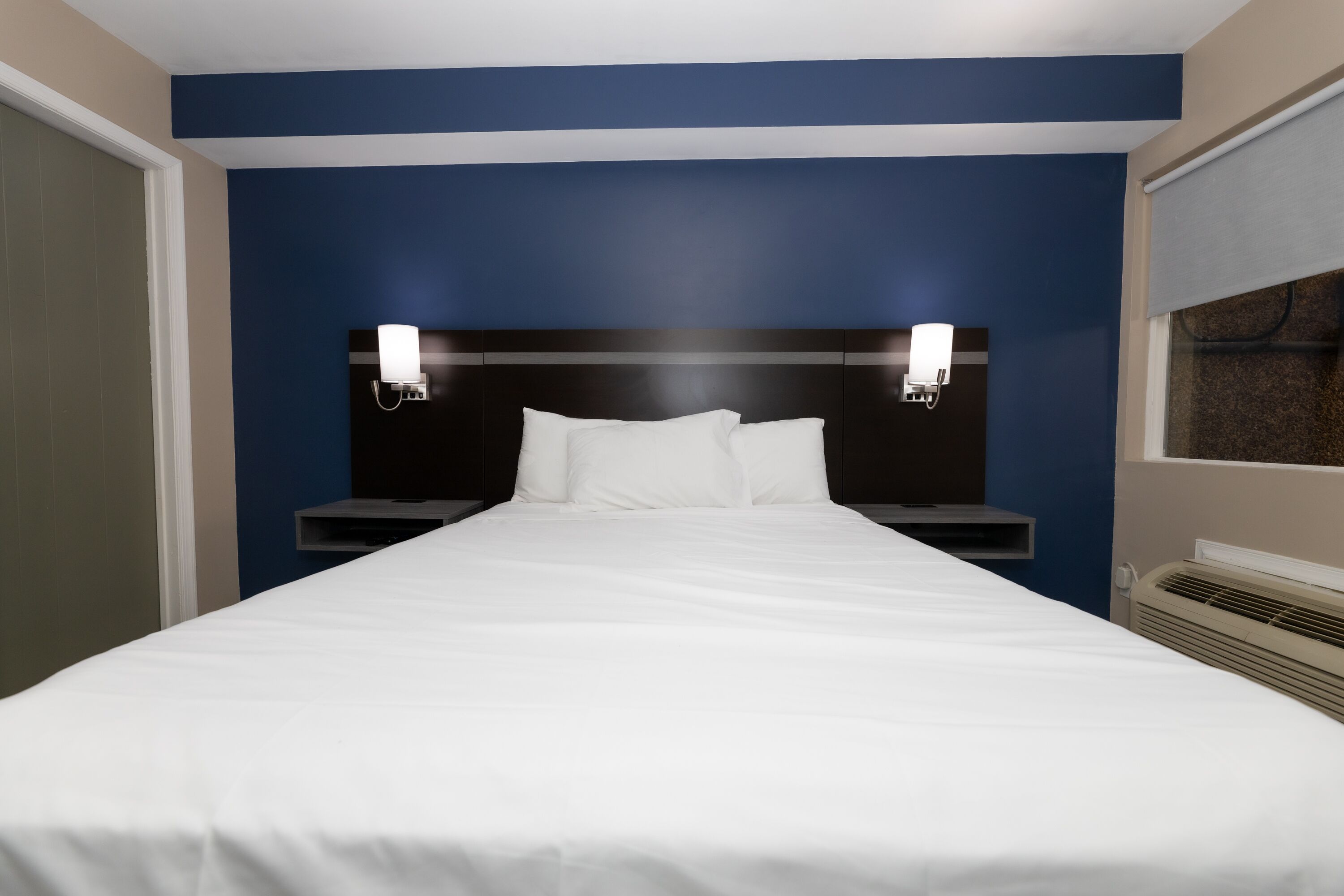 Quarto de hotel com uma cama grande, duas mesas de cabeceira, cabeceira com luminárias fixadas na parede e uma janela com persianas.