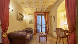 Uma sala de estar com sofá de couro, mesa de madeira e cadeira de vime. Há uma porta com janela e cortinas, e um teto com detalhes decorativos.