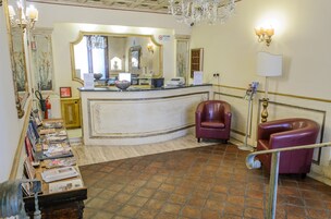 Um hall com uma mesa de recepção, duas poltronas de couro, um candelabro e piso de azulejos.