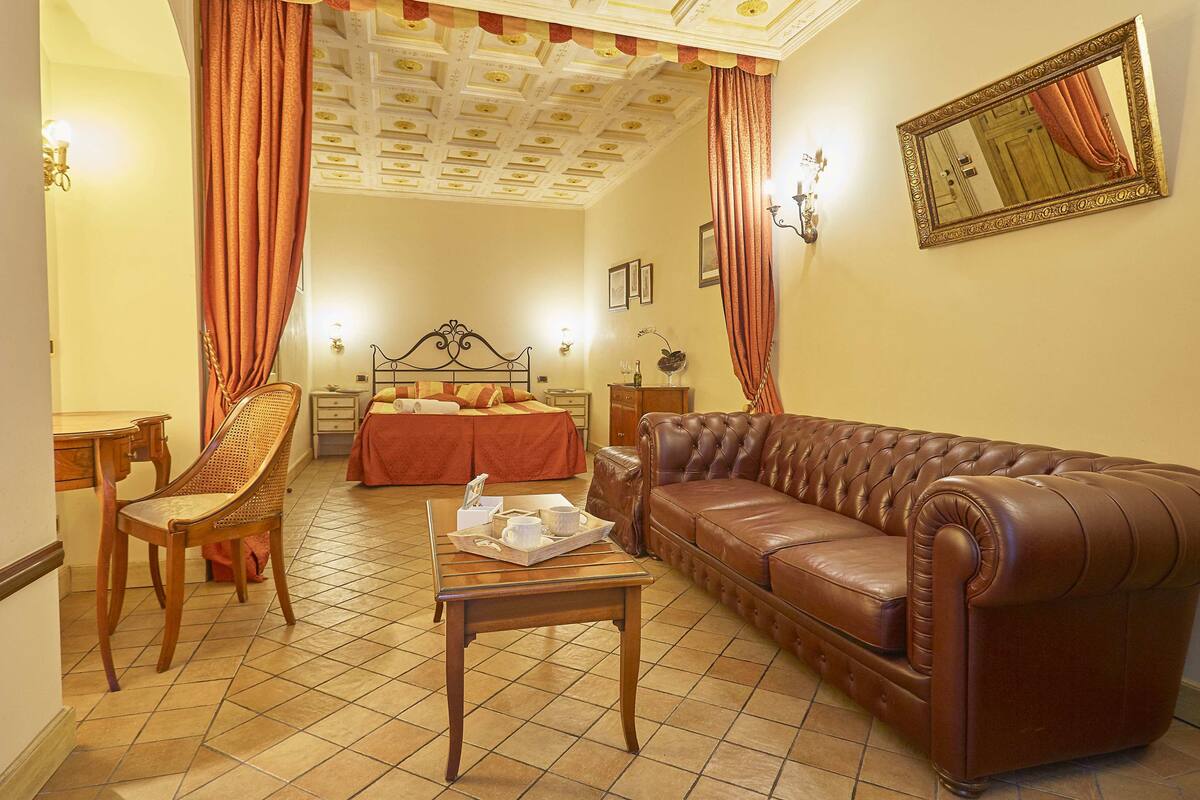 Quarto de hotel com uma cama, um sofá de couro, uma mesa de madeira, uma cadeira e um espelho na parede.