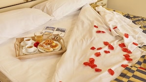 Cama bem arrumada com lençóis brancos, bandeja com itens de café da manhã como croissants, café e suco, e pétalas de rosas espalhadas.