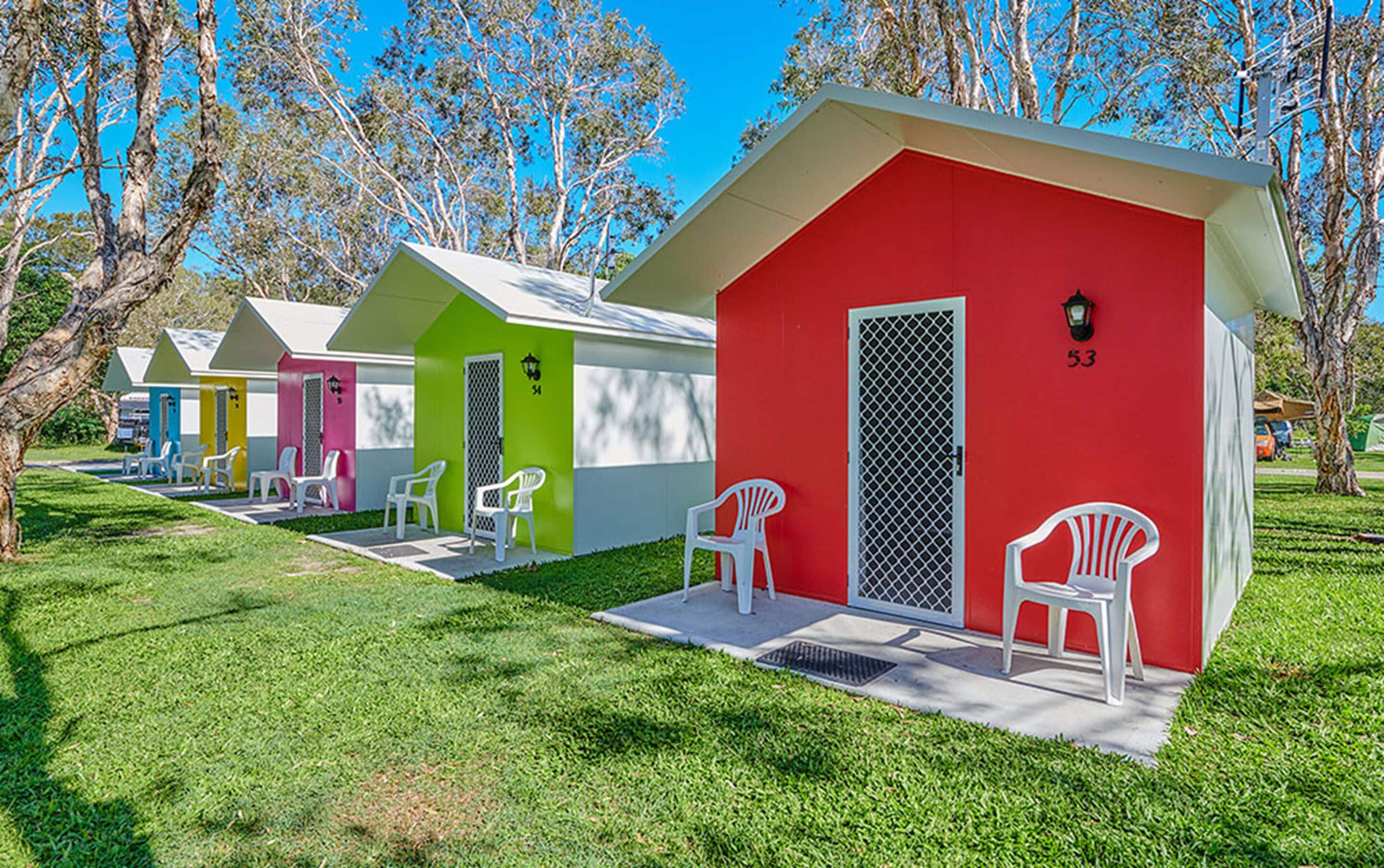 Uma fileira de cabanas coloridas, de um andar, com cadeiras de plástico brancas na parte externa, numeradas 53 e 54, situada em uma área gramada com árvores ao fundo.