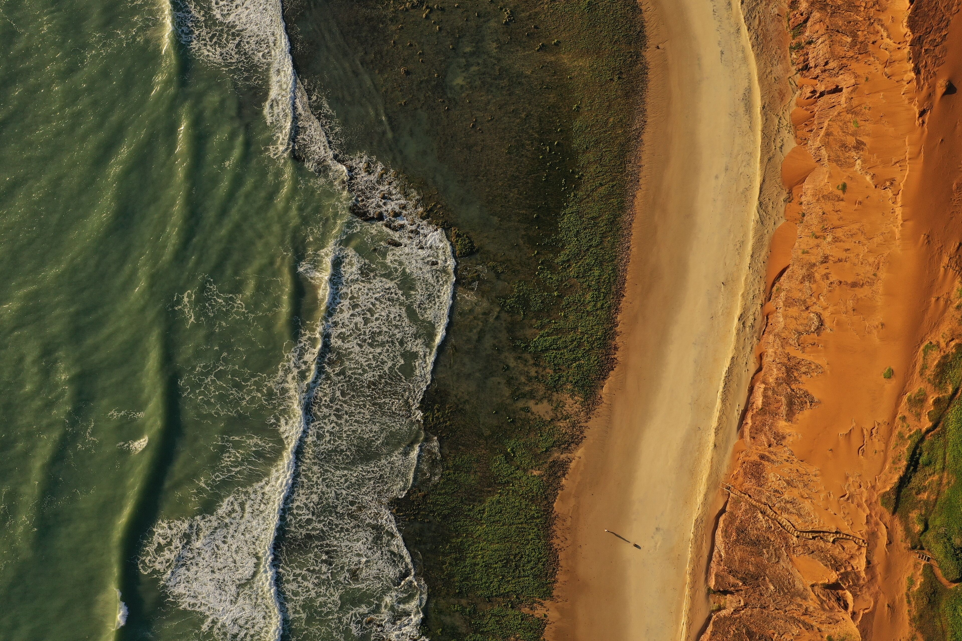 Vista aérea de uma praia com ondas quebrando na costa, uma faixa de areia e um penhasco rochoso.