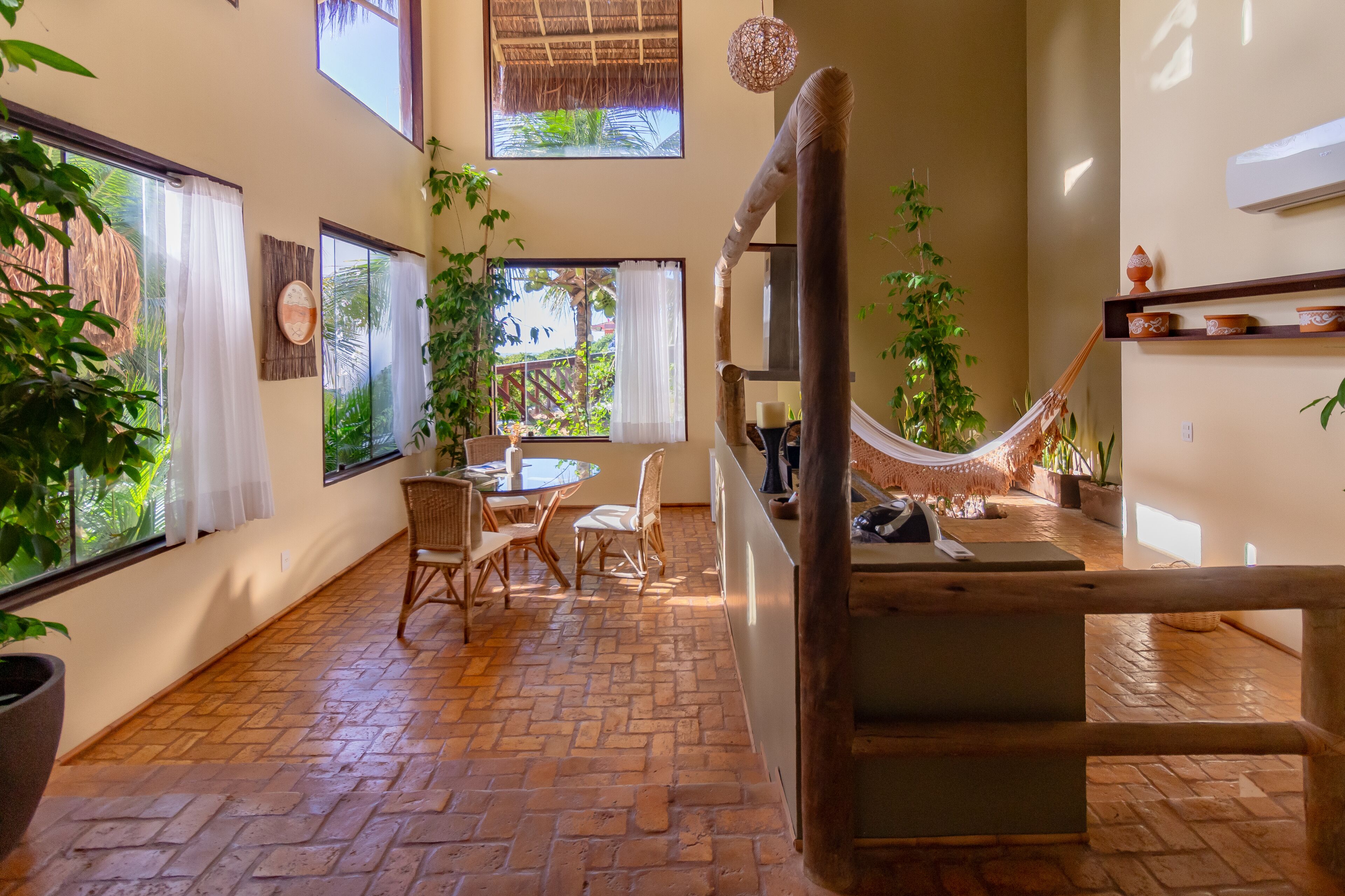 Um quarto espaçoso com piso de tijolos, grandes janelas com cortinas brancas, área de refeições com cadeiras, uma rede e plantas em vasos.