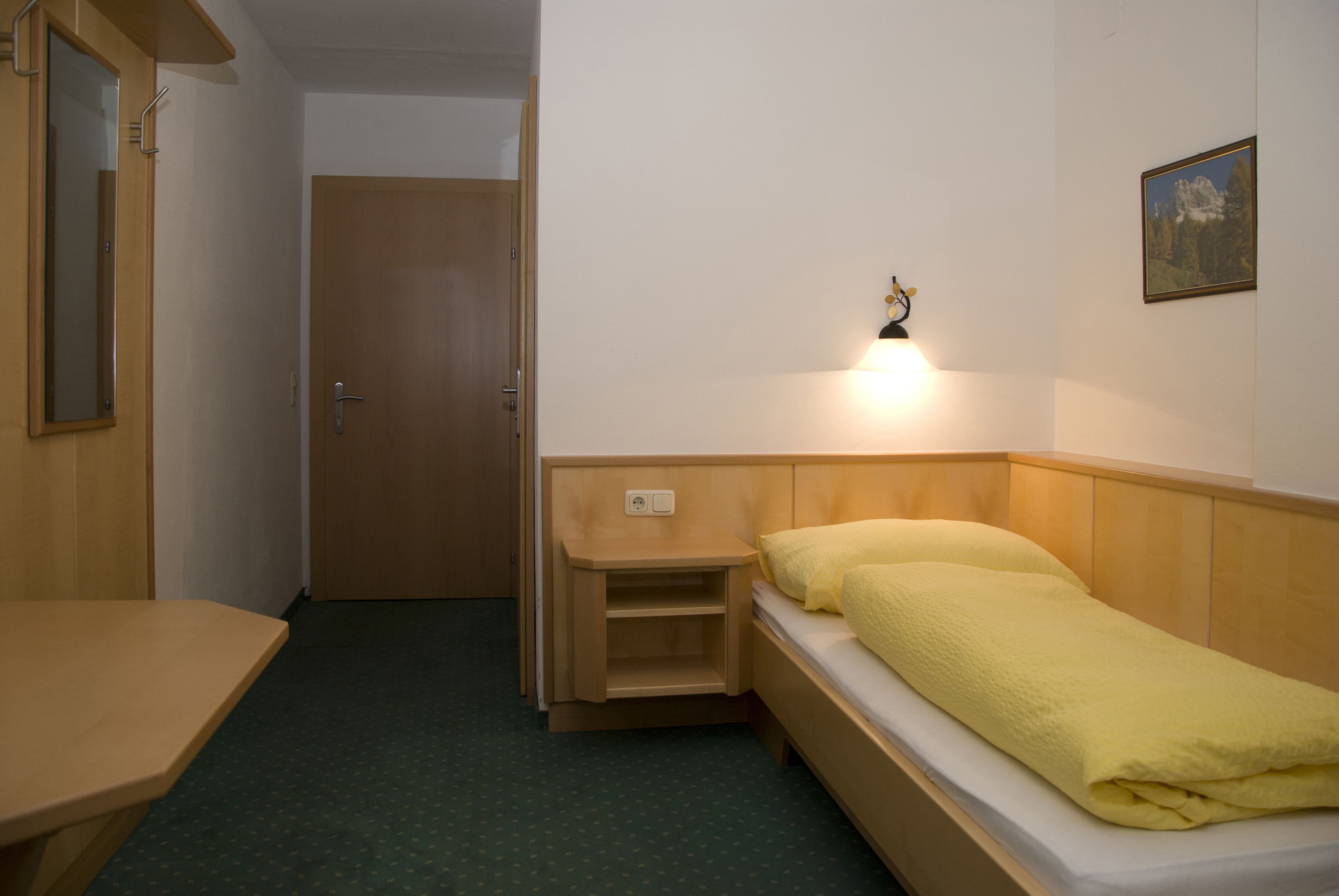 Quarto pequeno com cama de solteiro, cabeceira de madeira, mesa de cabeceira e luz acoplada na parede.