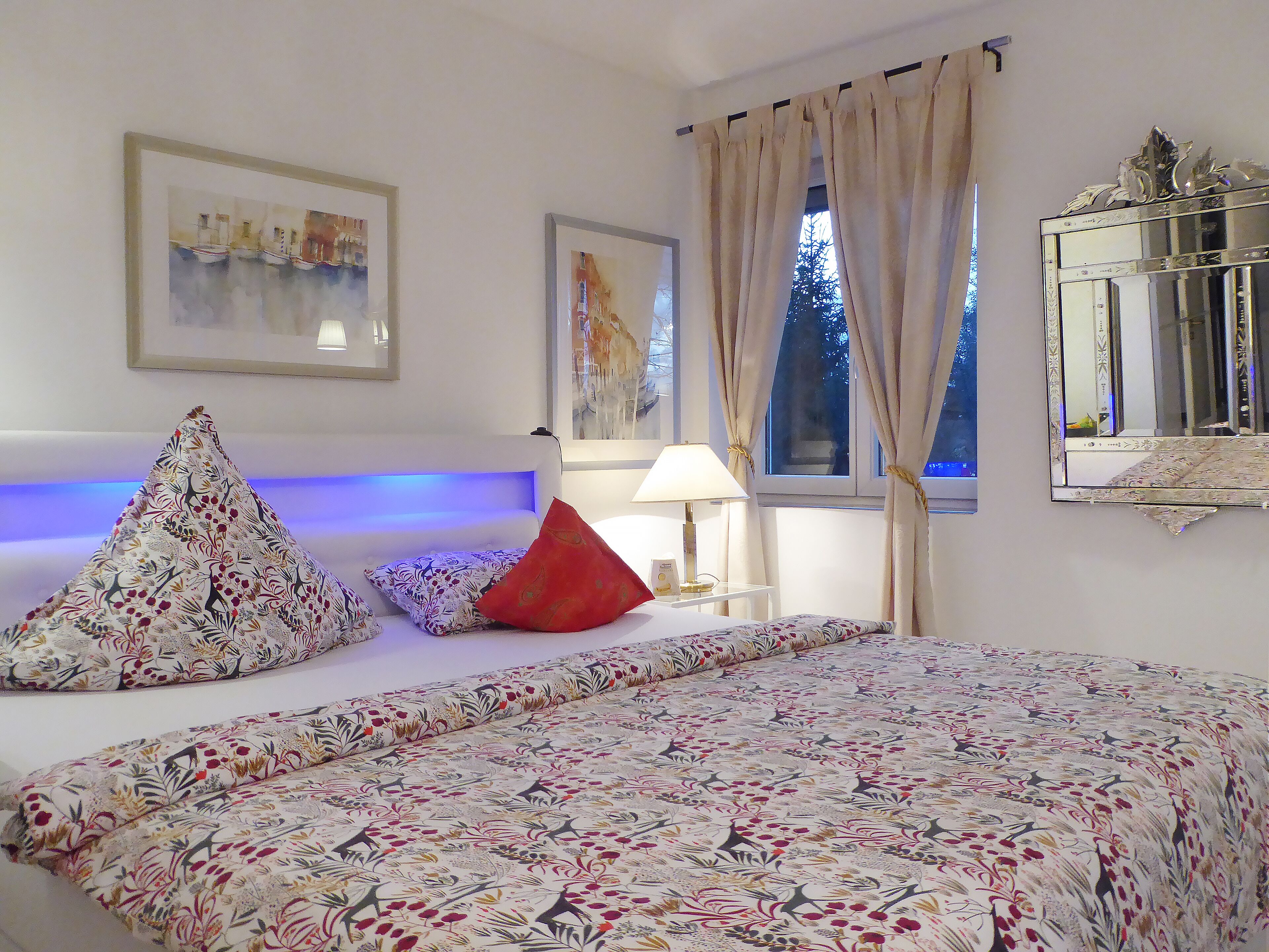 Um quarto com uma cama, uma mesa de cabeceira com abajur, uma janela com cortinas e duas obras de arte emolduradas na parede.