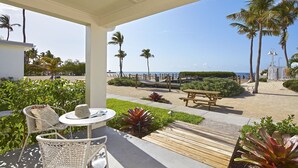 Uma área de estar coberta ao ar livre com cadeiras de vime e uma mesa, com vista para uma praia com palmeiras e um deck de madeira.