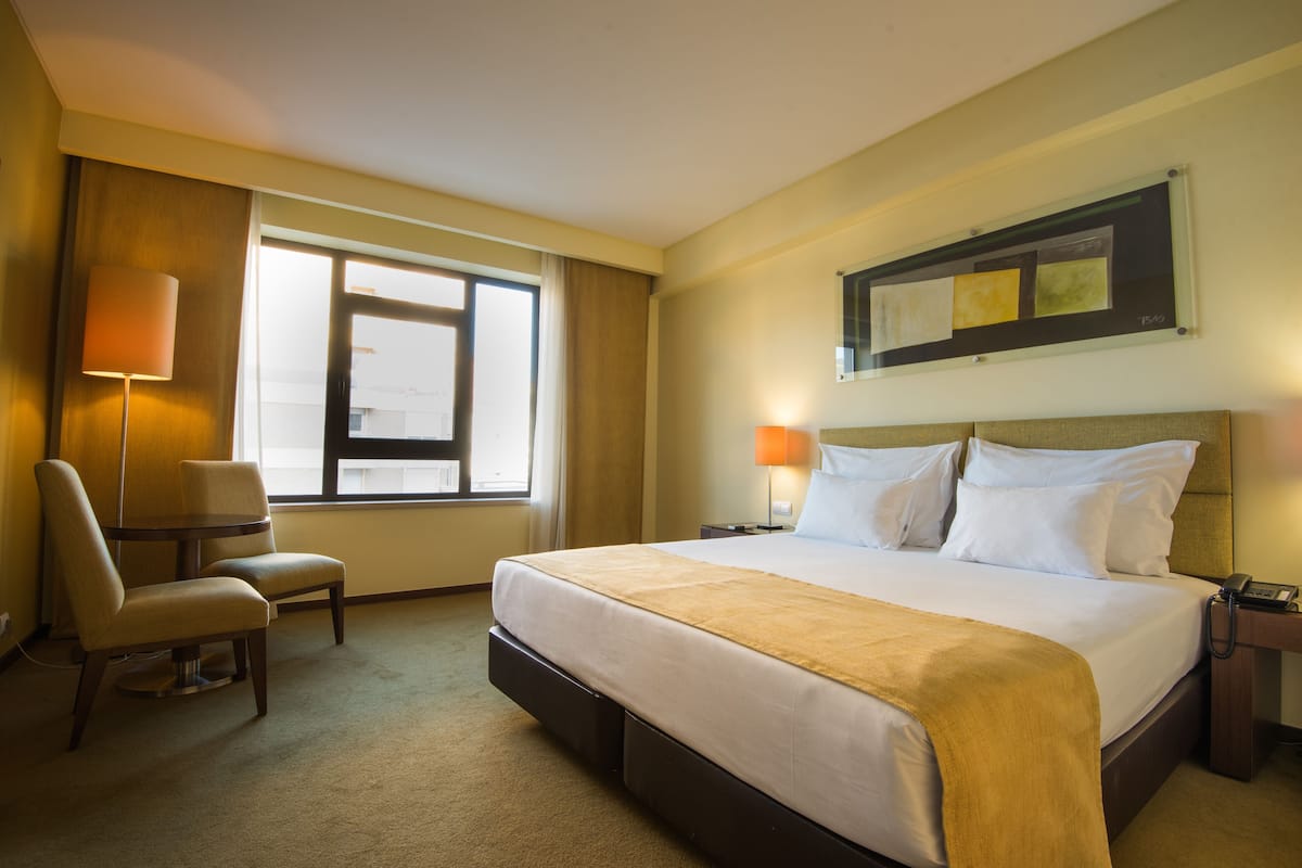 Quarto de hotel com uma cama grande, uma cadeira, uma escrivaninha e uma janela.