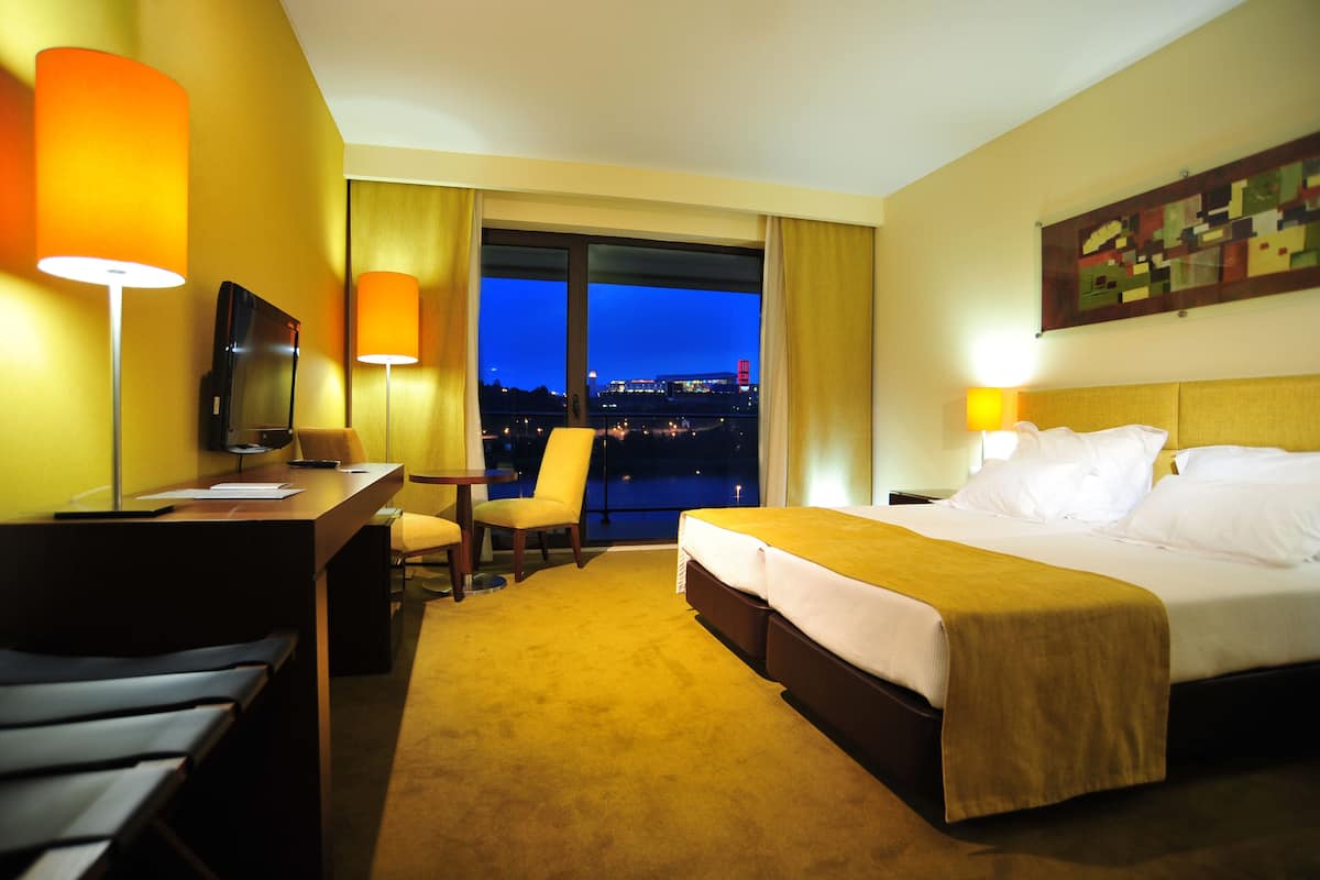 Quarto de hotel com cama, escrivaninha, cadeira e uma janela grande com vista para a cidade à noite.