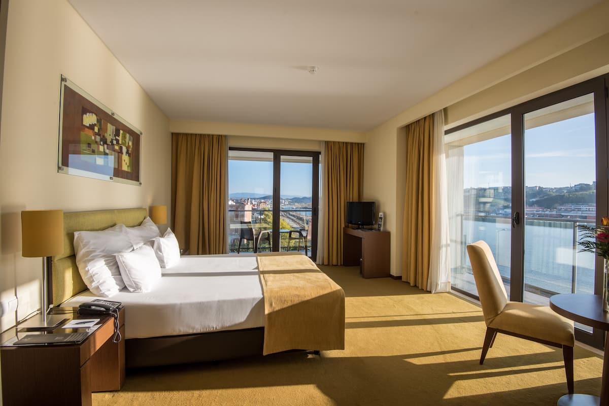 Quarto de hotel com cama, mesas de cabeceira, escrivaninha, cadeira, televisão e varanda com vista para a cidade.