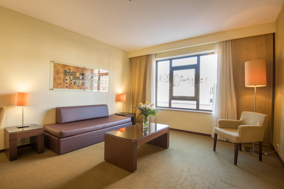 Quarto de hotel com sofá, mesa de centro, cadeira, luminária e janela com cortinas.