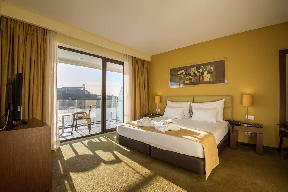 Quarto de hotel com uma cama grande, uma escrivaninha, uma cadeira, uma televisão e vista para a cidade pela janela.