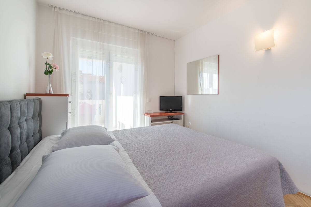 Quarto de hotel moderno com uma cama grande, televisão de tela plana, mesa de cabeceira e janela com cortinas finas.