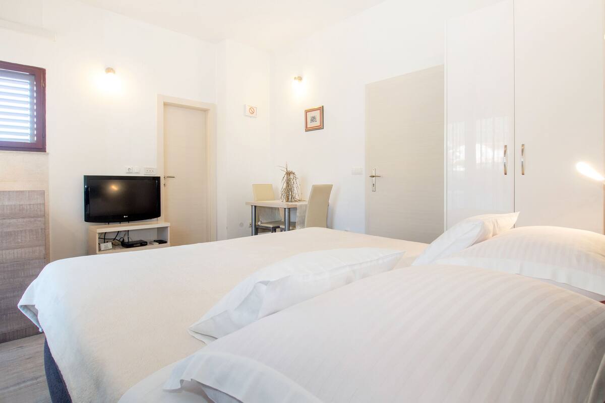 Quarto de hotel com uma cama grande, televisão sobre um suporte, uma mesinha de cabeceira com abajur e um guarda-roupa.