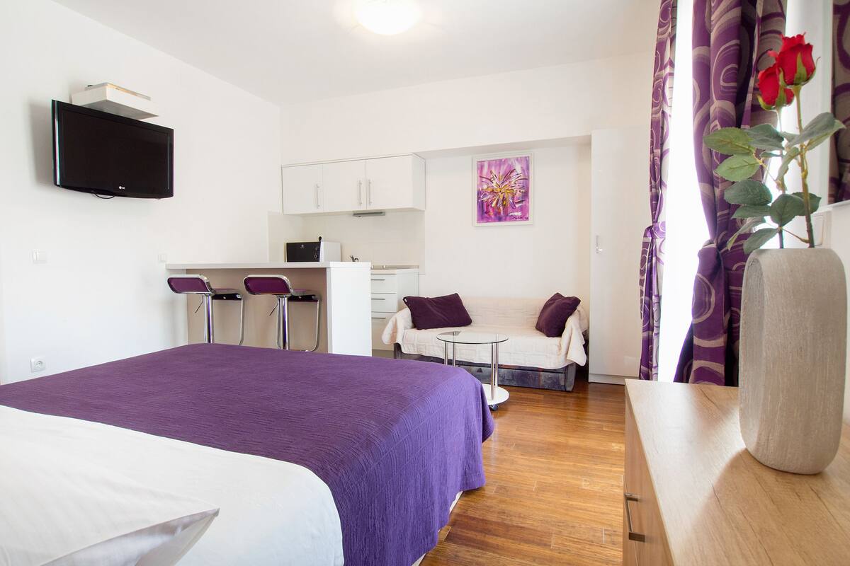 Quarto de hotel com cama, escrivaninha, cadeira, mesa pequena, televisão e um vaso com uma rosa.
