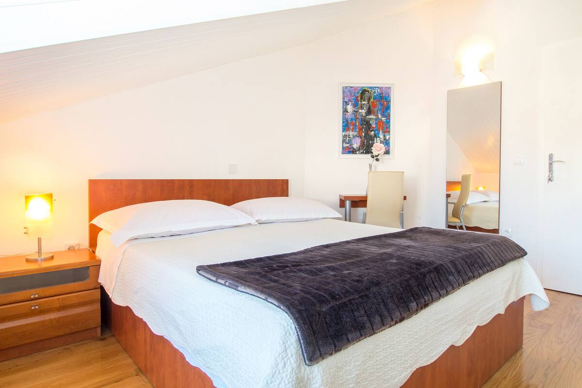Cama bem arrumada com edredom branco e manta escura, mesa de cabeceira com abajur, mesinha lateral com um vaso, e um quadro na parede.