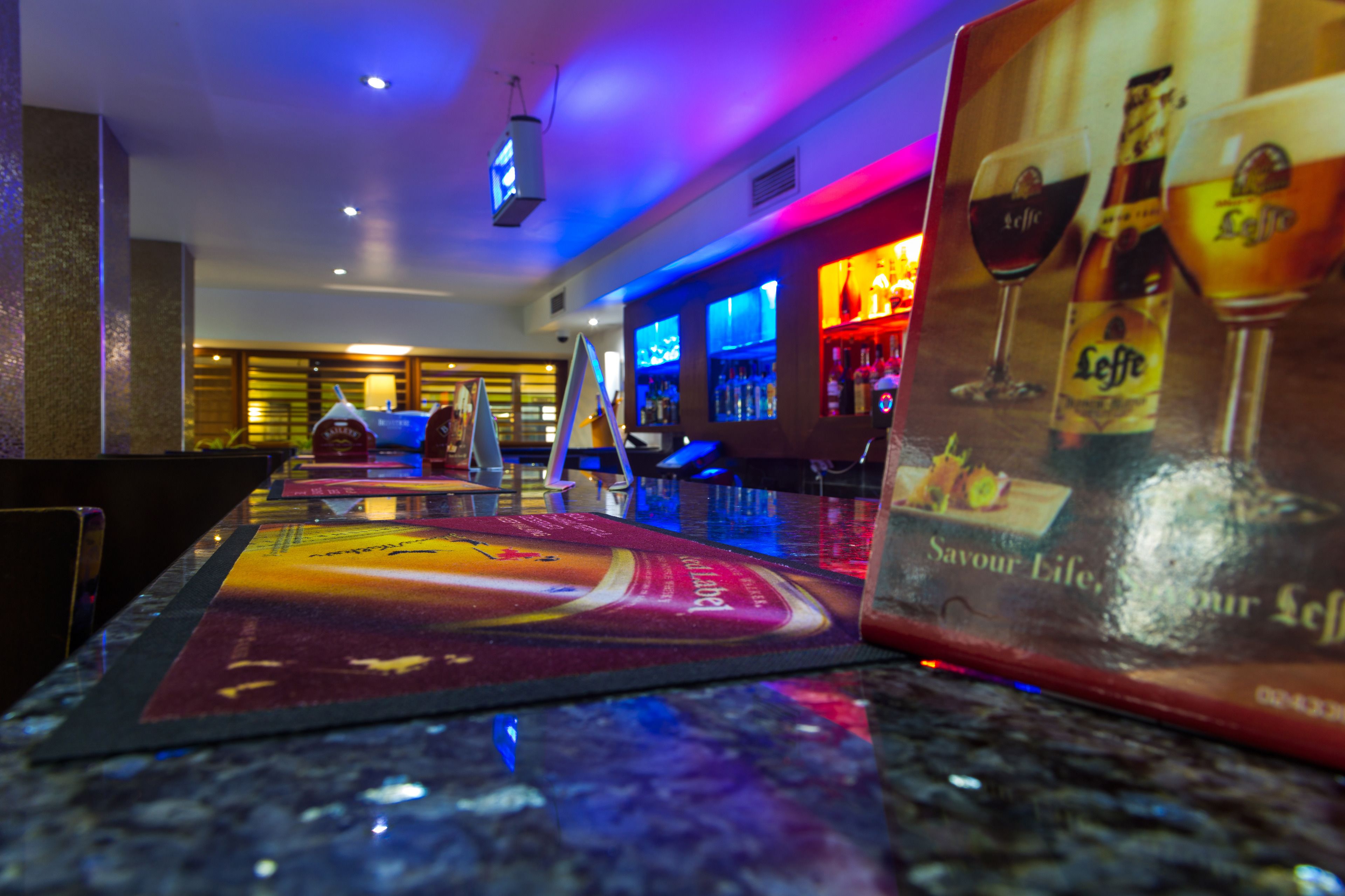 Uma área de bar com um cartaz anunciando cerveja Leffe, um balcão e um espaço de estar com mesas e cadeiras.