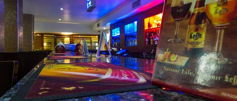 Uma área de bar com um cartaz anunciando cerveja Leffe, um balcão e um espaço de estar com mesas e cadeiras.