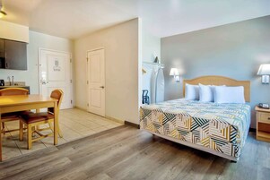 Quarto de hotel com uma cama, uma escrivaninha e uma cadeira. Há uma porta e uma prateleira fixada na parede com objetos.